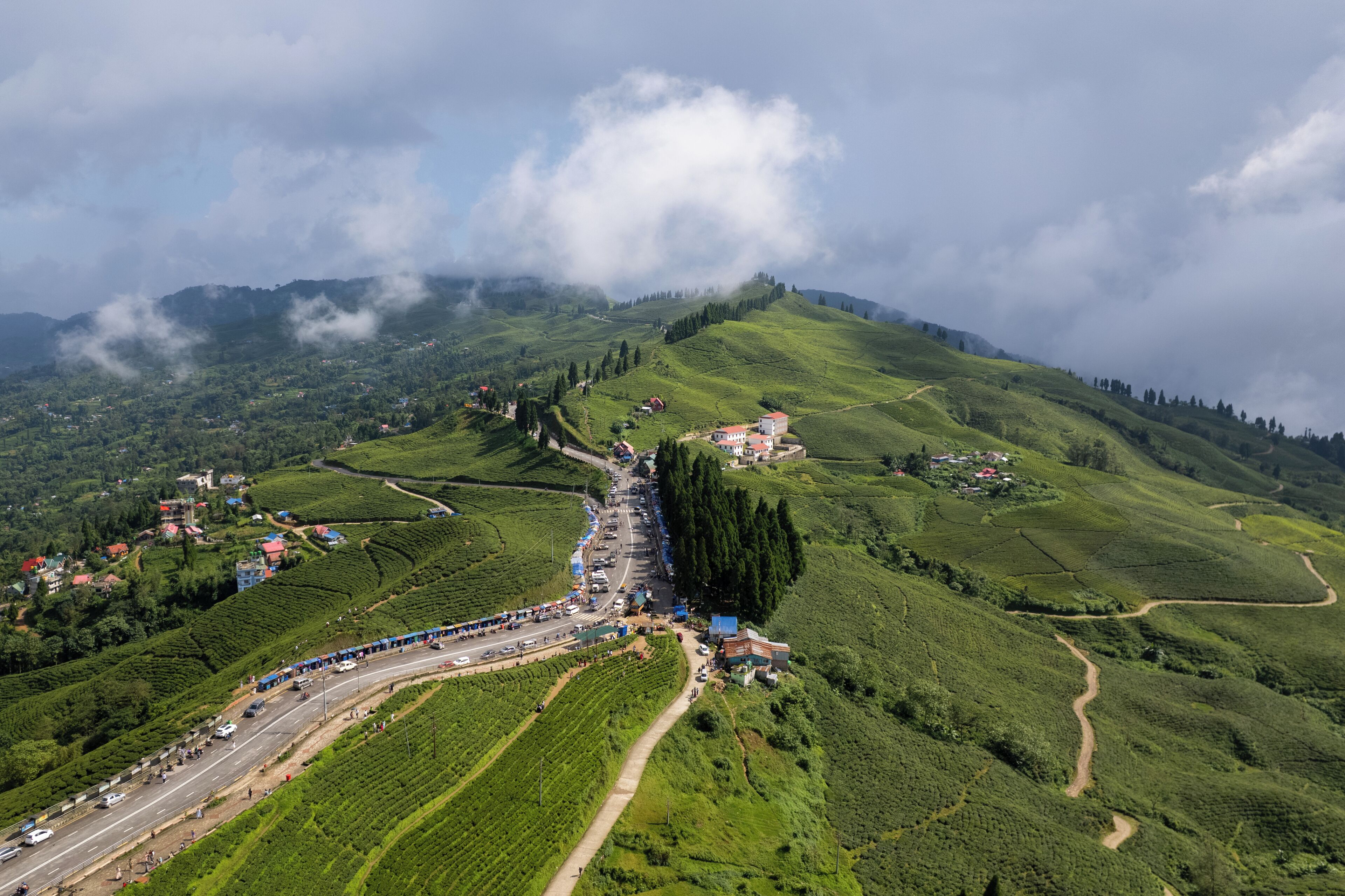 Paisagem acidentada com uma estrada serpenteando por campos em socalcos, alguns edifícios e uma grande formação de nuvens.