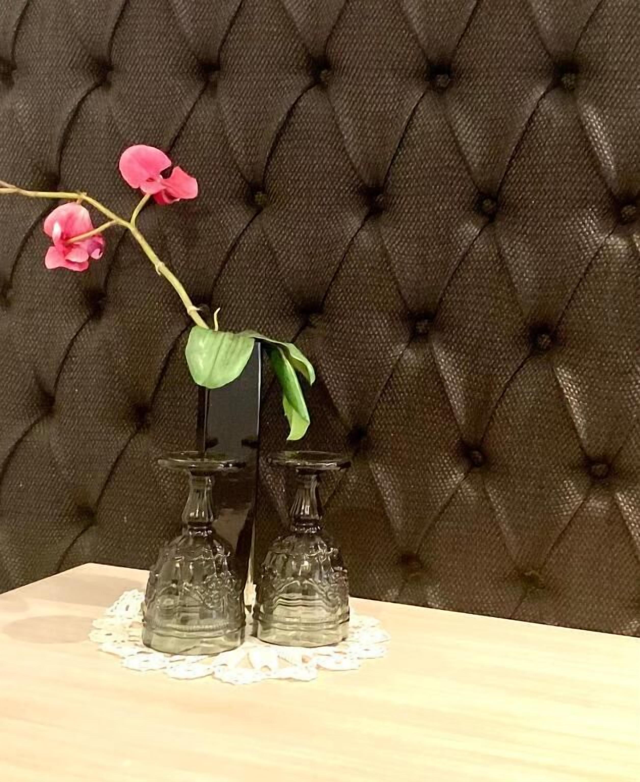 Dois vasos de vidro com flores cor-de-rosa sobre uma mesa de madeira, com uma parede de couro acolchoado ao fundo.