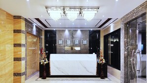 Uma área de recepção de hotel com um balcão de mármore, relógios na parede e um candelabro.