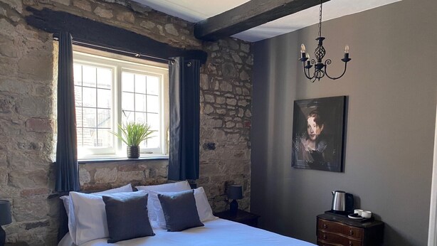 Quarto com parede de pedra, uma cama grande com roupa de cama branca e azul escura, uma planta em vaso perto da janela, um quadro na parede e uma mesa de cabeceira com abajur.