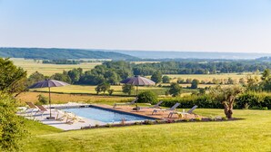 Área de piscina com espreguiçadeiras e guarda-sóis, cercada por paisagem verdejante e colinas ao longe.