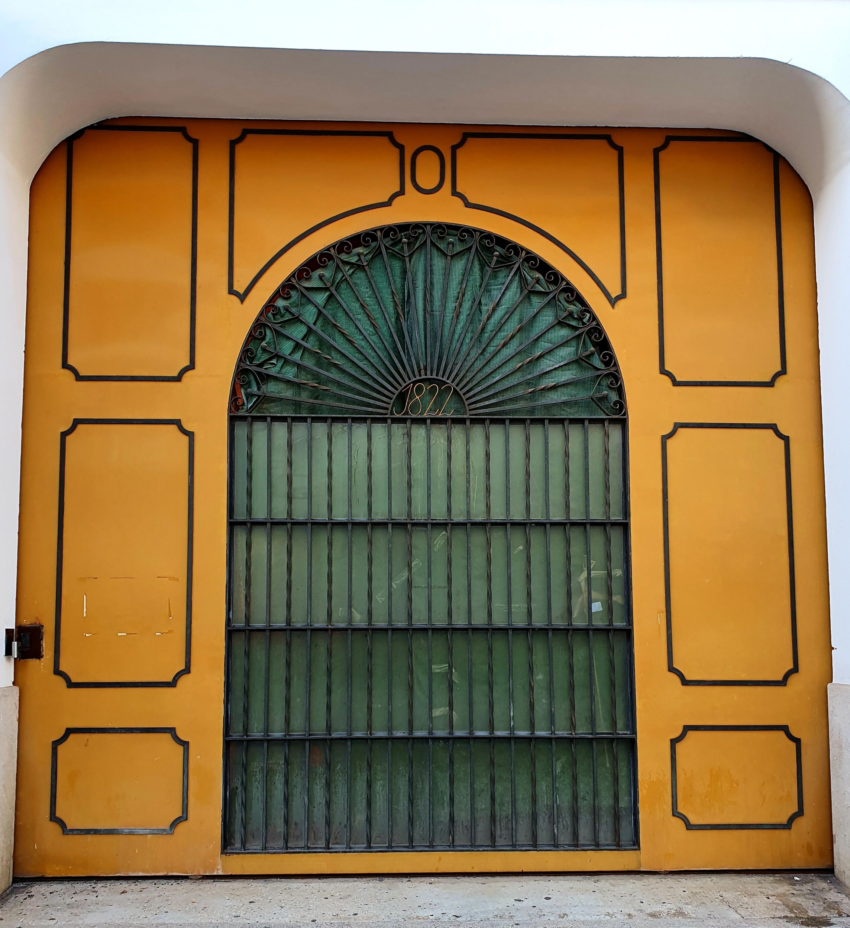 Uma porta amarela com um portão de metal verde e um janelão decorativo em formato de leque acima.