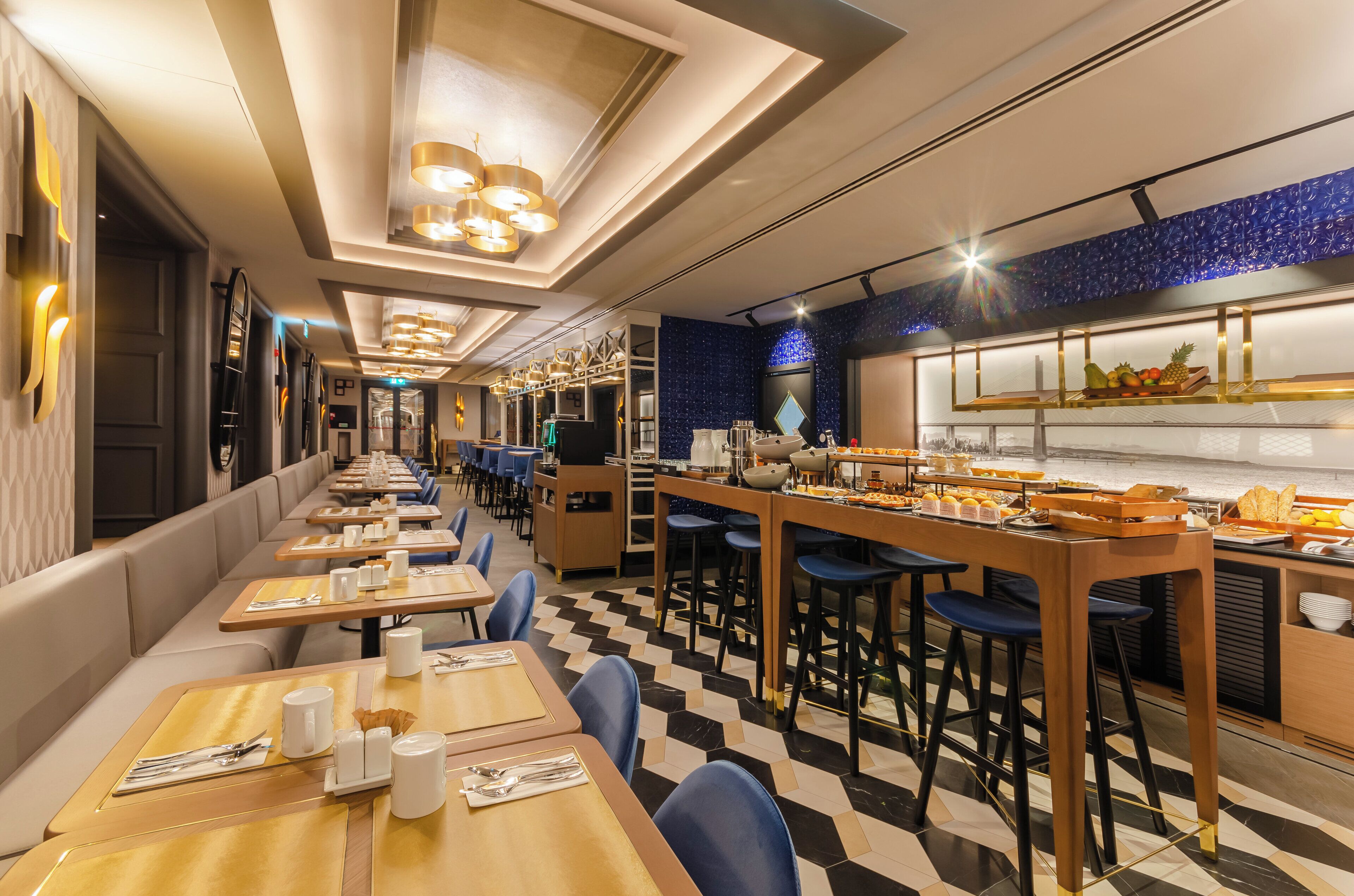 Restaurante com mesas compridas, cadeiras azuis e um buffet com comida e bebidas.