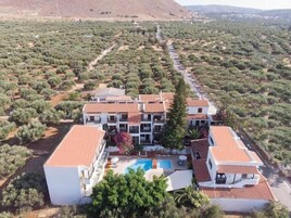 Um hotel com vários andares, piscina, rodeado por oliveiras e montanhas ao fundo.