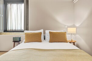 Uma cama bem arrumada com almofadas brancas e douradas, um abajur e uma pequena mesa com um relógio.