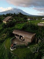 Uma casa com telhado de palha, um pátio de pedra e área de estar ao ar livre, rodeada por vegetação exuberante e uma montanha ao fundo.