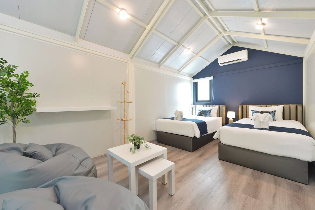 Quarto de hotel moderno com clarabóia, duas camas, uma mesinha e uma planta.