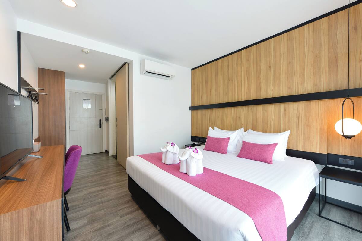 Quarto de hotel moderno com uma cama grande, uma escrivaninha e uma cadeira. Apresenta uma parede revestida em madeira e uma luminária no teto.
