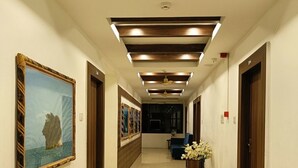 Um corredor de hotel com piso polido, portas de madeira, obras de arte emolduradas nas paredes e uma área de recepção com sofá azul e flores brancas.