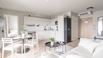 Uma cozinha moderna com armários brancos, uma sala de jantar com mesa e cadeiras redondas, e uma sala de estar com sofá e mesinha lateral.