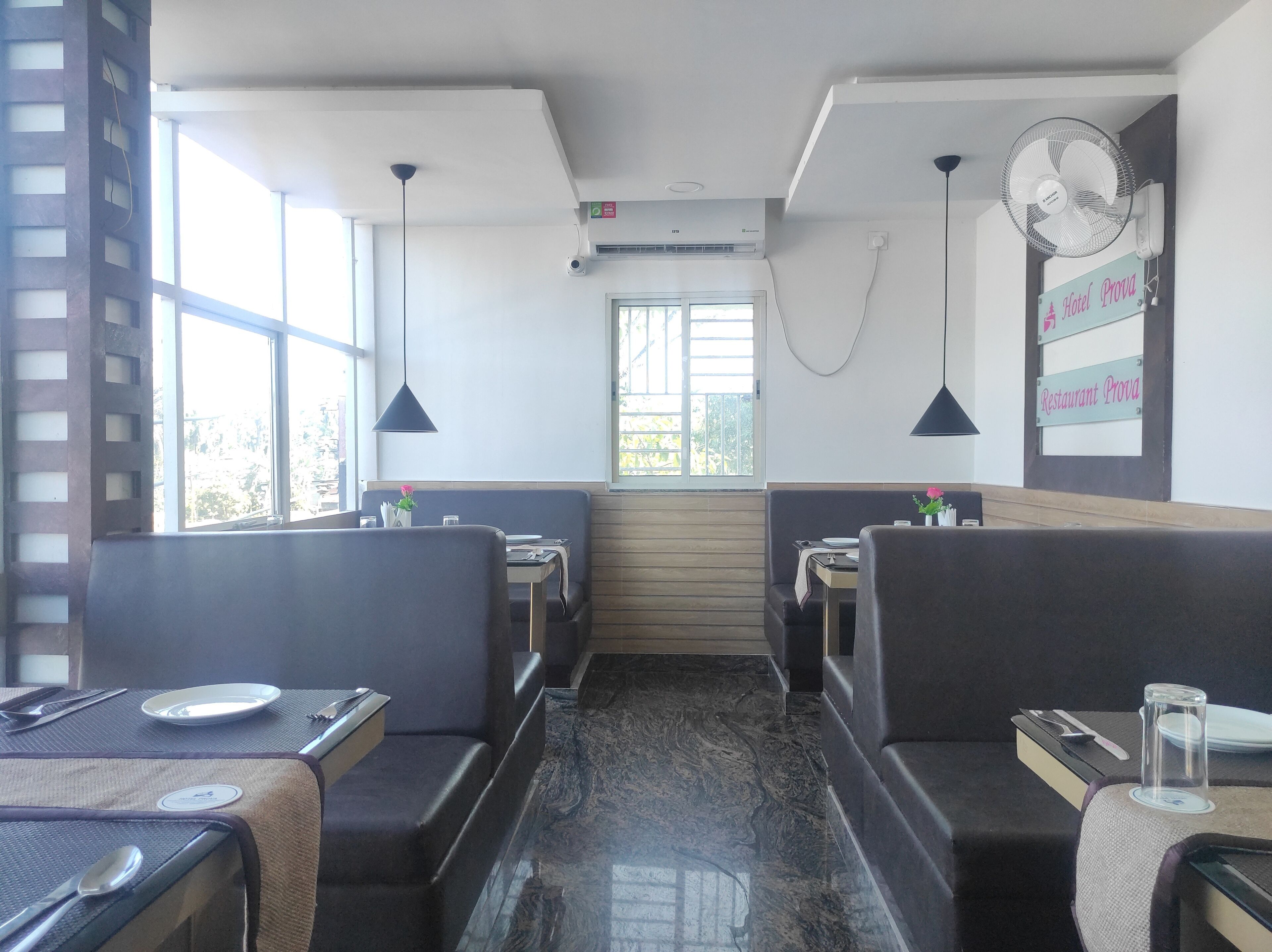 Restaurante com assentos em cabines, janela central, luminárias pendentes e um ventilador na parede.