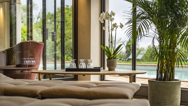 Quarto de hotel moderno com uma janela grande, um sofá confortável, uma mesa pequena com uma orquídea em vaso e vista para uma piscina.
