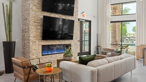Um lobby moderno com parede de tijolos, duas grandes TVs de tela plana, área de estar com sofá e poltronas, uma pequena mesa com uma planta e uma janela grande com vista para o exterior.