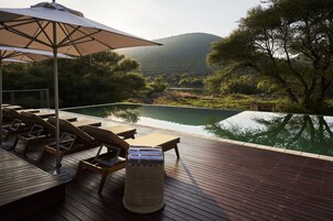Um deck de madeira com cadeiras de lounge e uma mesa, com vista para águas calmas e uma colina arborizada.