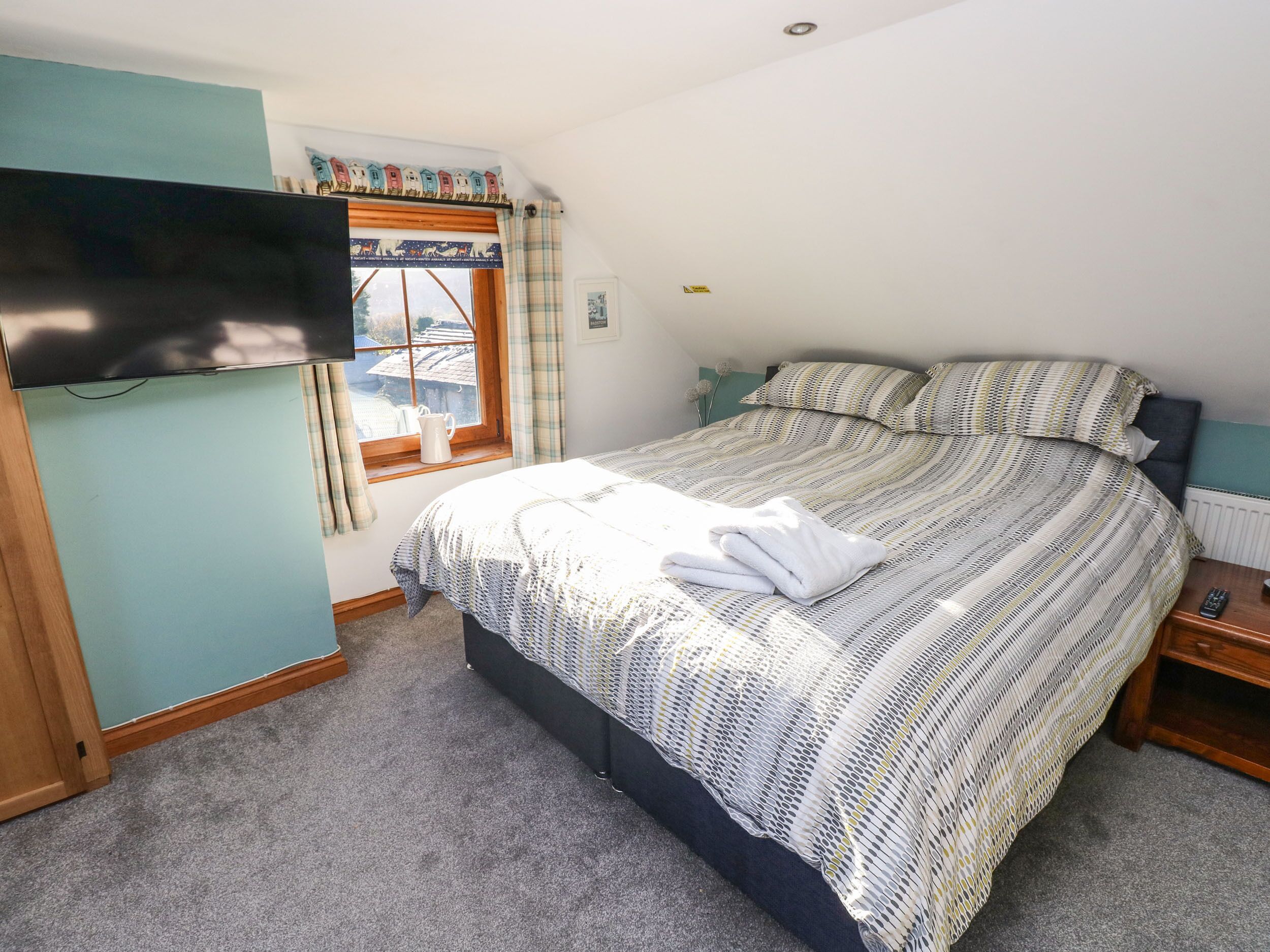 Quarto com cama, televisão fixada na parede, janela com cortinas e mesa de cabeceira.
