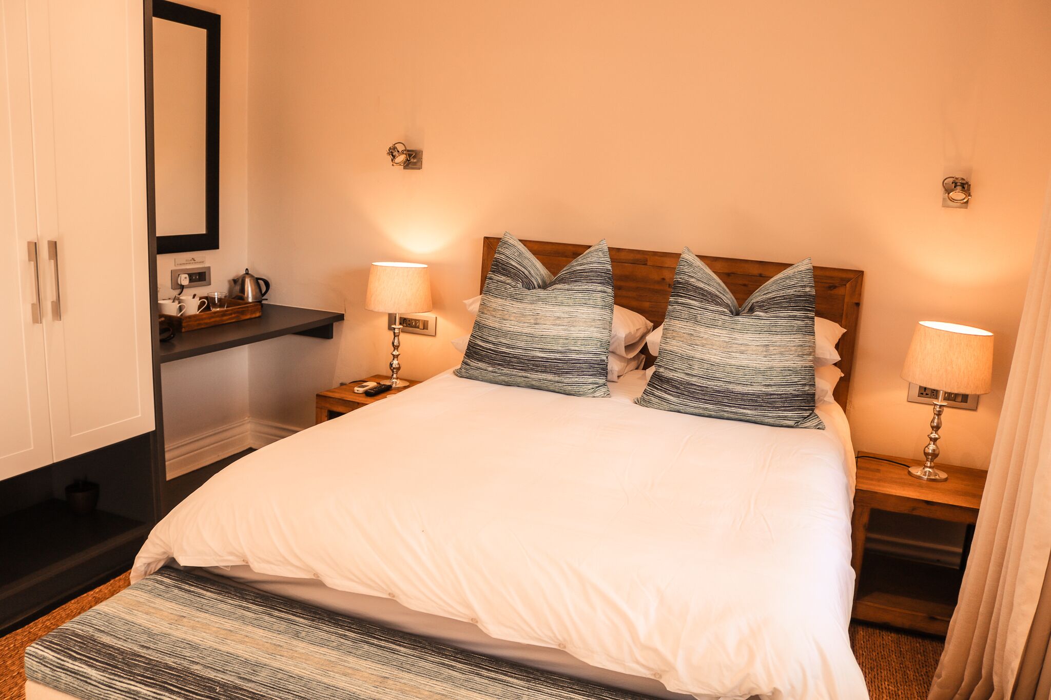 Quarto bem arrumado com uma cama grande, duas mesas de cabeceira com abajures, uma cabeceira de madeira e uma pequena prateleira com chaleira e xícaras.