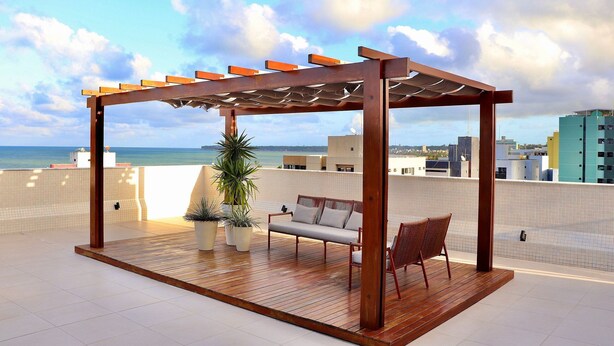 Uma pérgola de madeira com sofá e cadeiras em um terraço no telhado, com vista para o mar e edifícios.