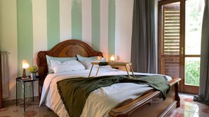Quarto com cama de madeira, duas mesas de cabeceira com luminárias, janela com persianas e vista para o verde lá fora.