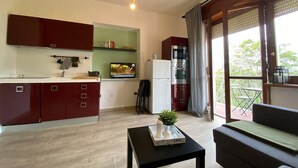Uma cozinha moderna com armários vermelho escuro, uma geladeira branca e um forno embutido. Há uma área de jantar com uma mesa preta e um sofá. Uma televisão está fixada acima do balcão da cozinha.