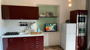 Uma cozinha compacta com fogão a gás, pia branca e forno embutido. Há uma televisão fixada na parede e uma geladeira à direita.