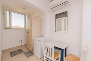 Quarto pequeno e limpo com uma escrivaninha branca, duas cadeiras brancas, um frigorífico branco compacto, um guarda-roupa de madeira e uma janela com vista para uma montanha.