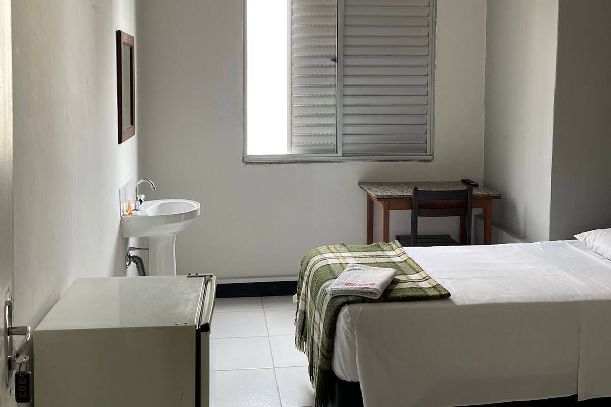 Quarto de hotel com cama, uma mesinha, uma cadeira, pia e janela com persianas.