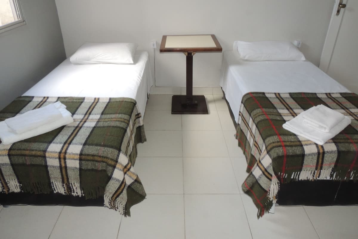 Quarto com duas camas de solteiro, cada uma com um cobertor xadrez e uma toalha dobrada. Há uma pequena mesa com uma luminária entre as camas.