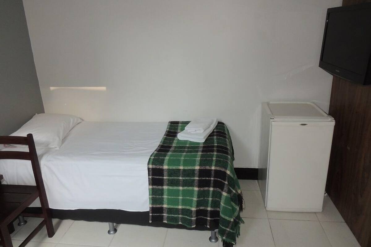 Quarto pequeno e simples com cama, uma cadeira, televisão e um armário branco.