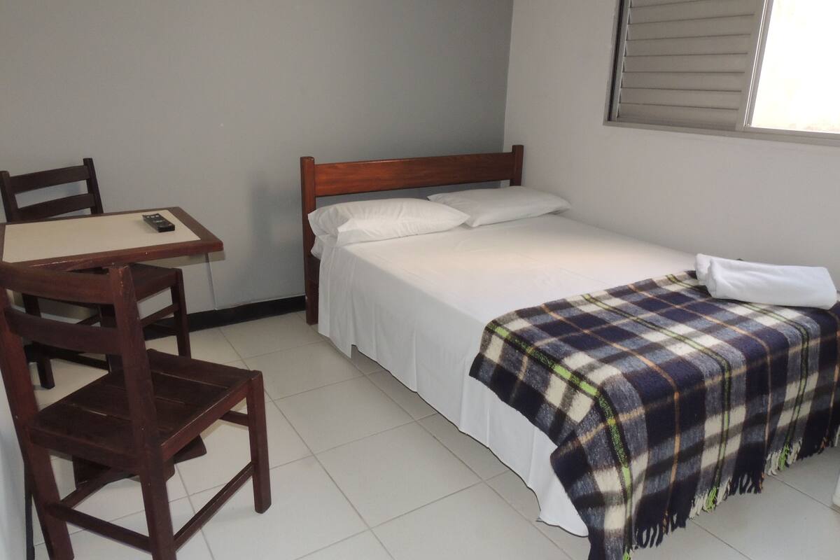Quarto pequeno e arrumado, com uma cama de solteiro, uma escrivaninha de madeira e uma cadeira.