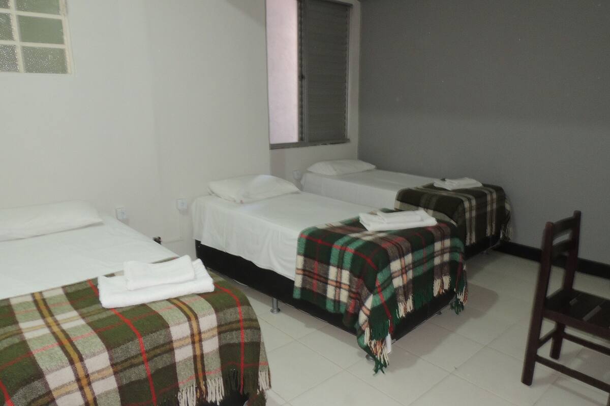Quarto de hotel com três camas de solteiro, cada uma com colchão branco e manta xadrez. Há uma janela na parede esquerda e uma cadeira no canto.