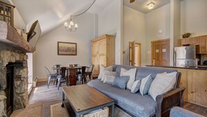 Uma sala de estar acolhedora com lareira de pedra, um sofá cinza, armários de madeira e uma área de cozinha.