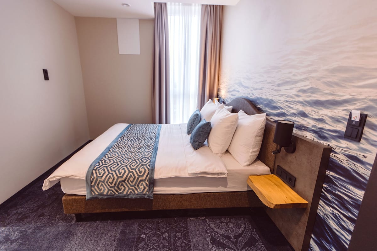 Quarto de hotel com cama, mesa de cabeceira, luminária na parede e vista para o mar pela janela.