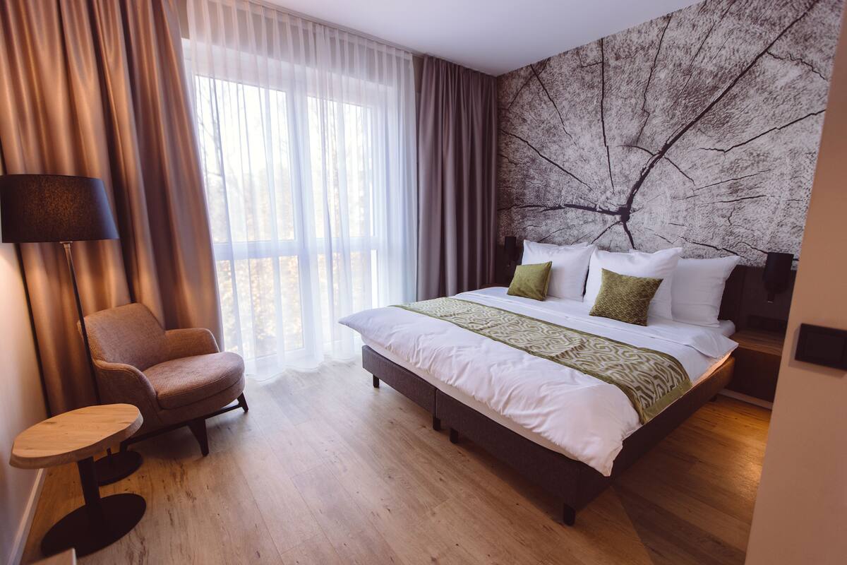 Quarto de hotel com uma cama grande, uma cadeira, uma mesa pequena e vista para o exterior através de cortinas translúcidas.
