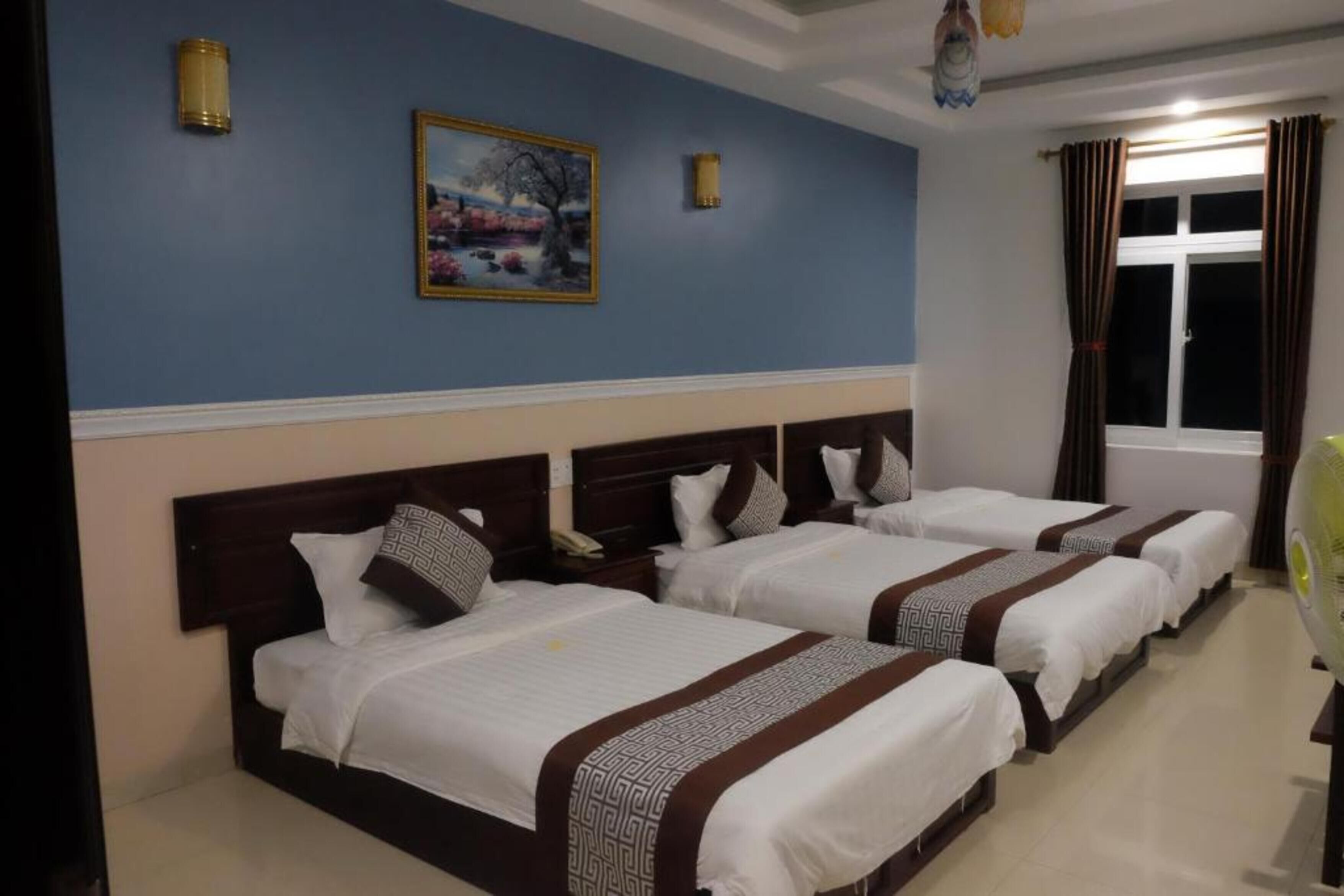 Quarto de hotel com três camas alinhadas contra uma parede azul, cada uma com colcha e almofadas estampadas. Há um quadro acima das camas e uma janela com cortinas à direita.
