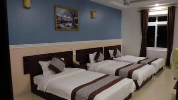 Quarto de hotel com três camas alinhadas contra uma parede azul, cada uma com colcha e almofadas estampadas. Há um quadro acima das camas e uma janela com cortinas à direita.
