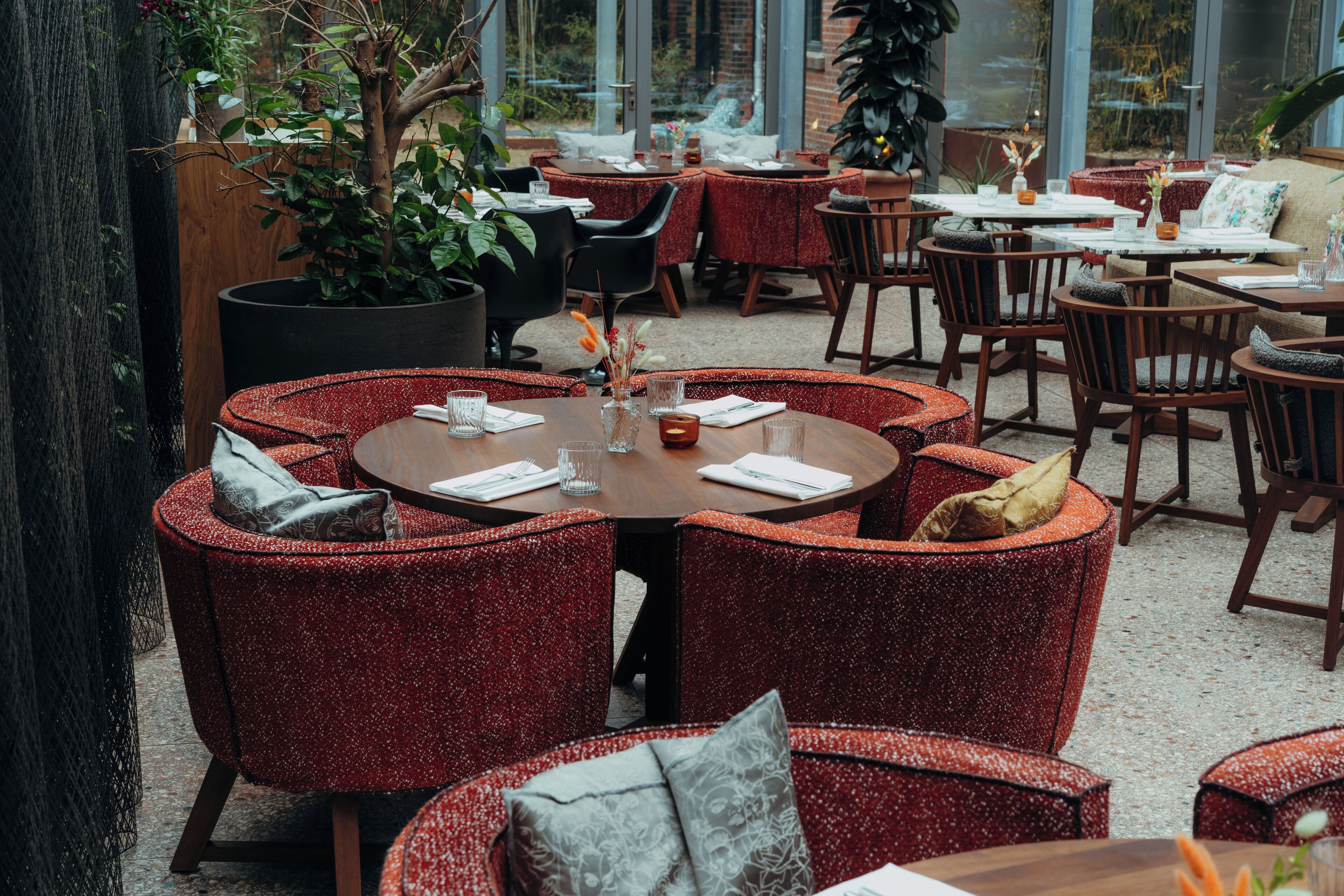 Restaurante com mesas redondas e cadeiras estofadas vermelhas, rodeado por plantas em vasos e grandes janelas.