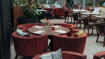 Restaurante com mesas redondas e cadeiras estofadas vermelhas, rodeado por plantas em vasos e grandes janelas.