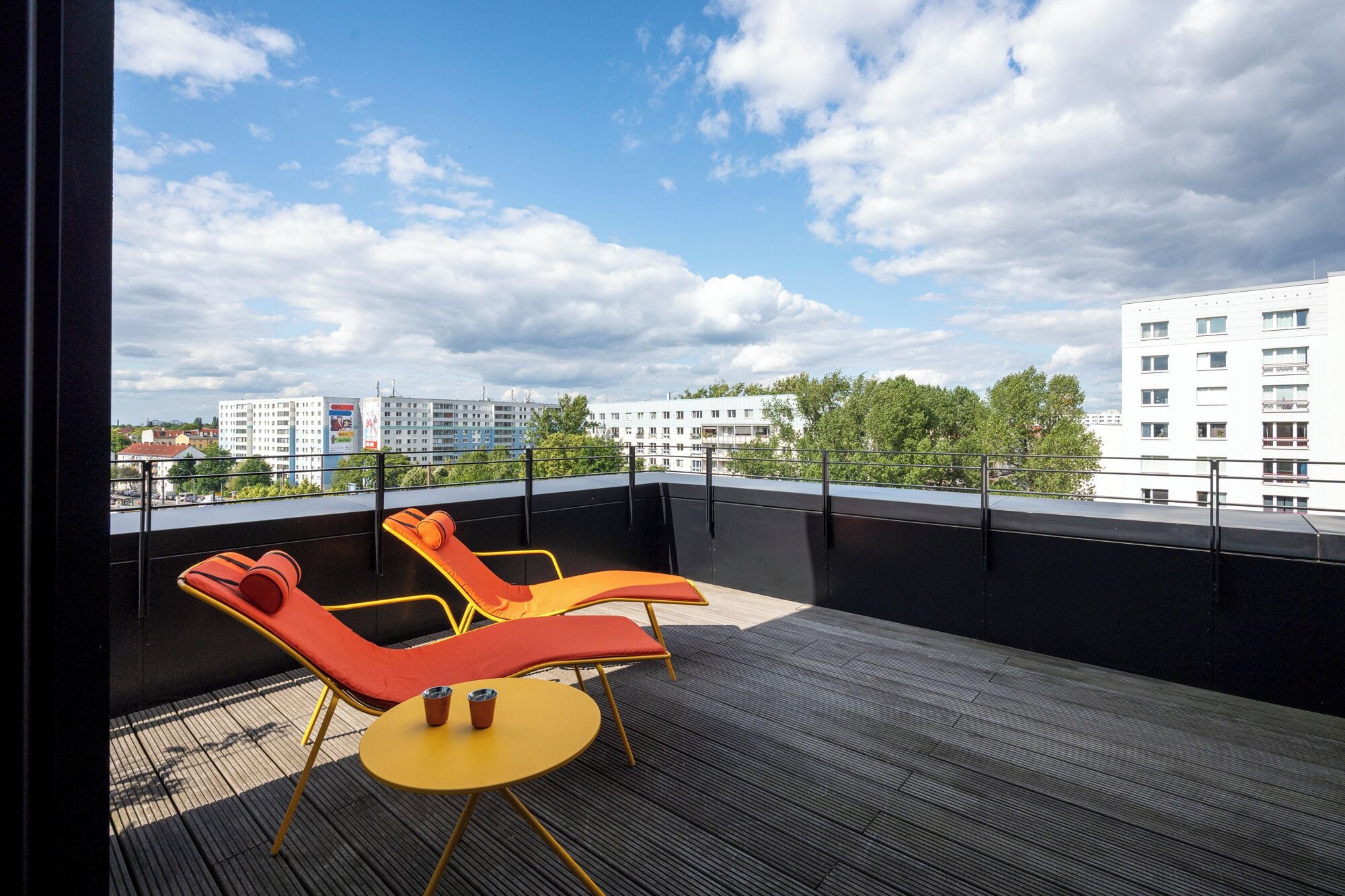 Varanda com duas cadeiras de lounge laranja e uma mesa redonda pequena, com vista para um horizonte urbano com edifícios modernos.