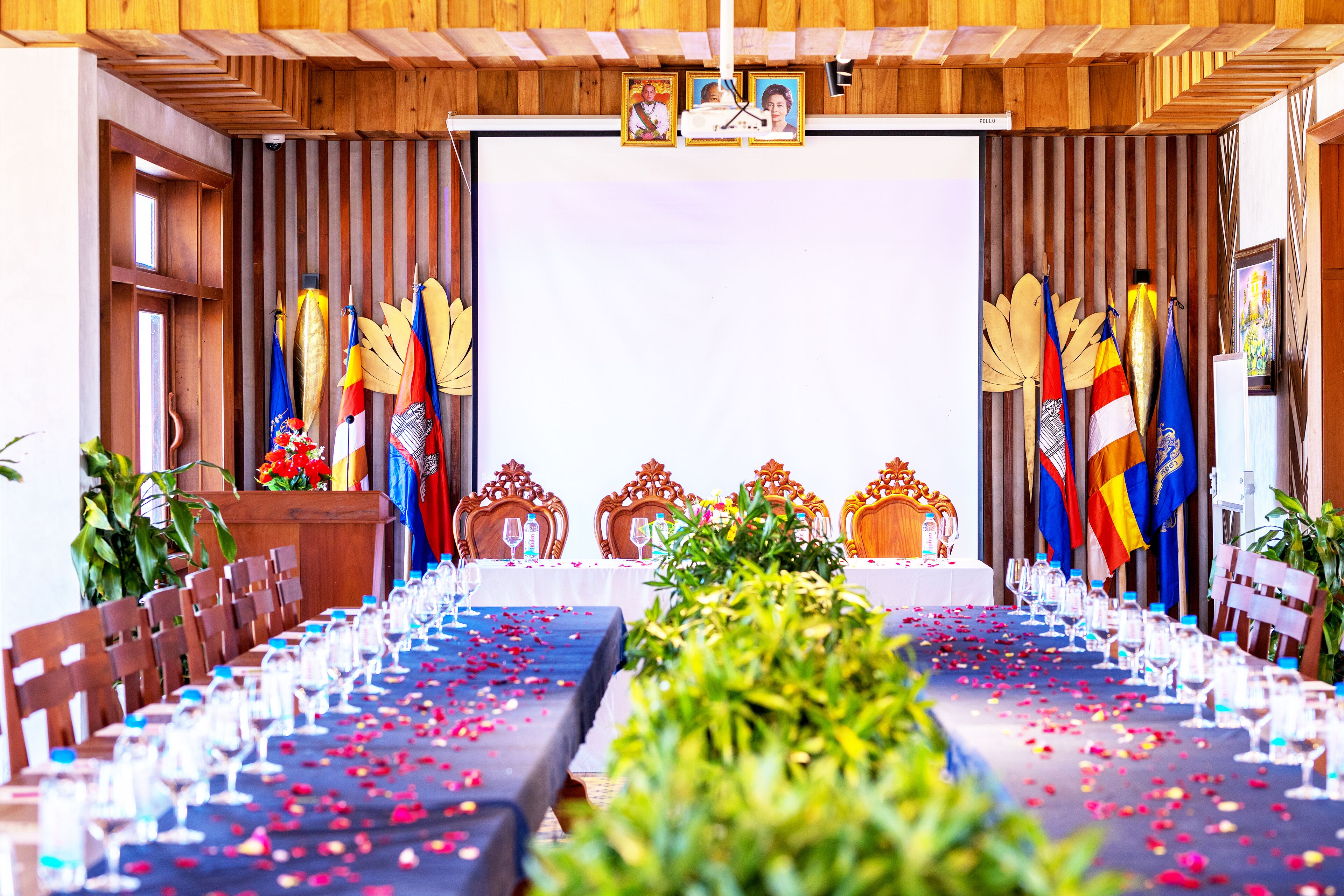 Uma longa mesa de banquete posta para um evento formal, com bandeiras e cadeiras de madeira de cada lado.