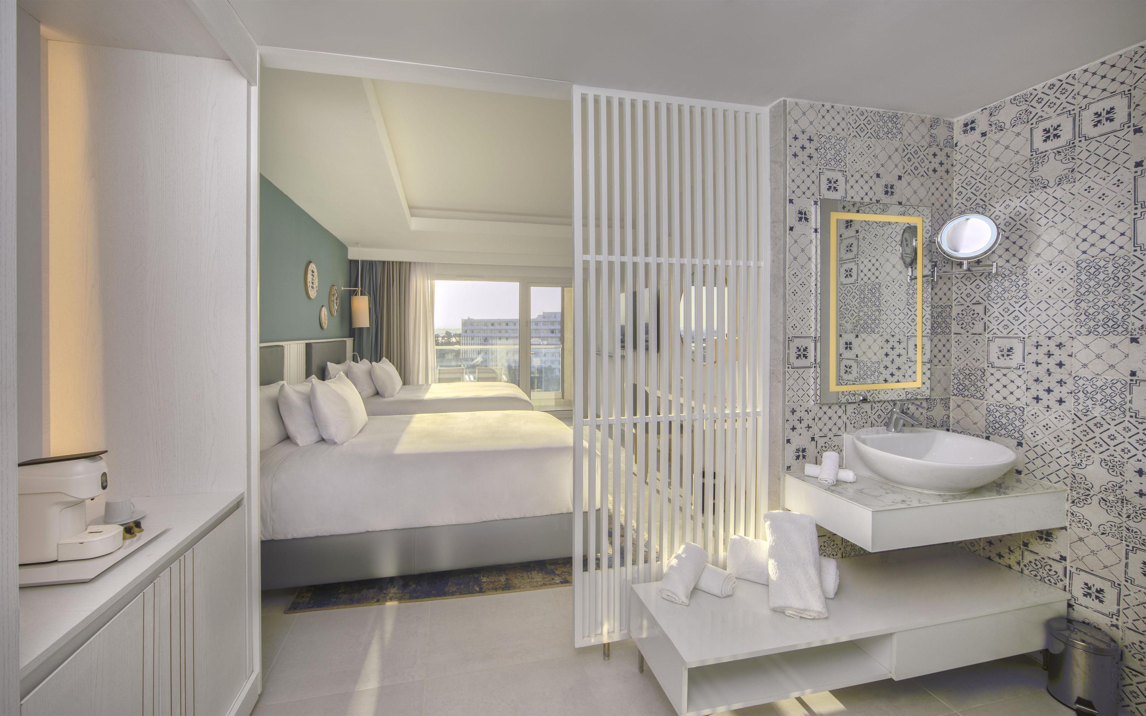 Quarto de hotel moderno com cama, banheiro com pia e espelho, e vista para a cidade.