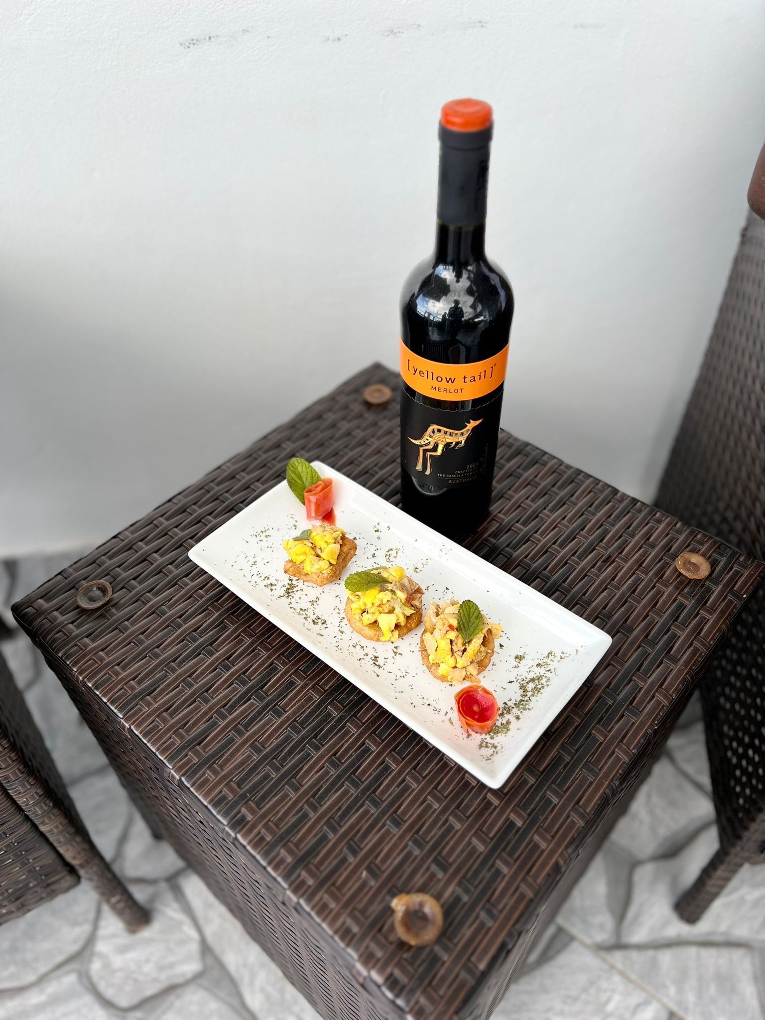 Uma garrafa de vinho Merlot [yellow tail] sobre uma mesa de palha com um prato de aperitivos brancos.