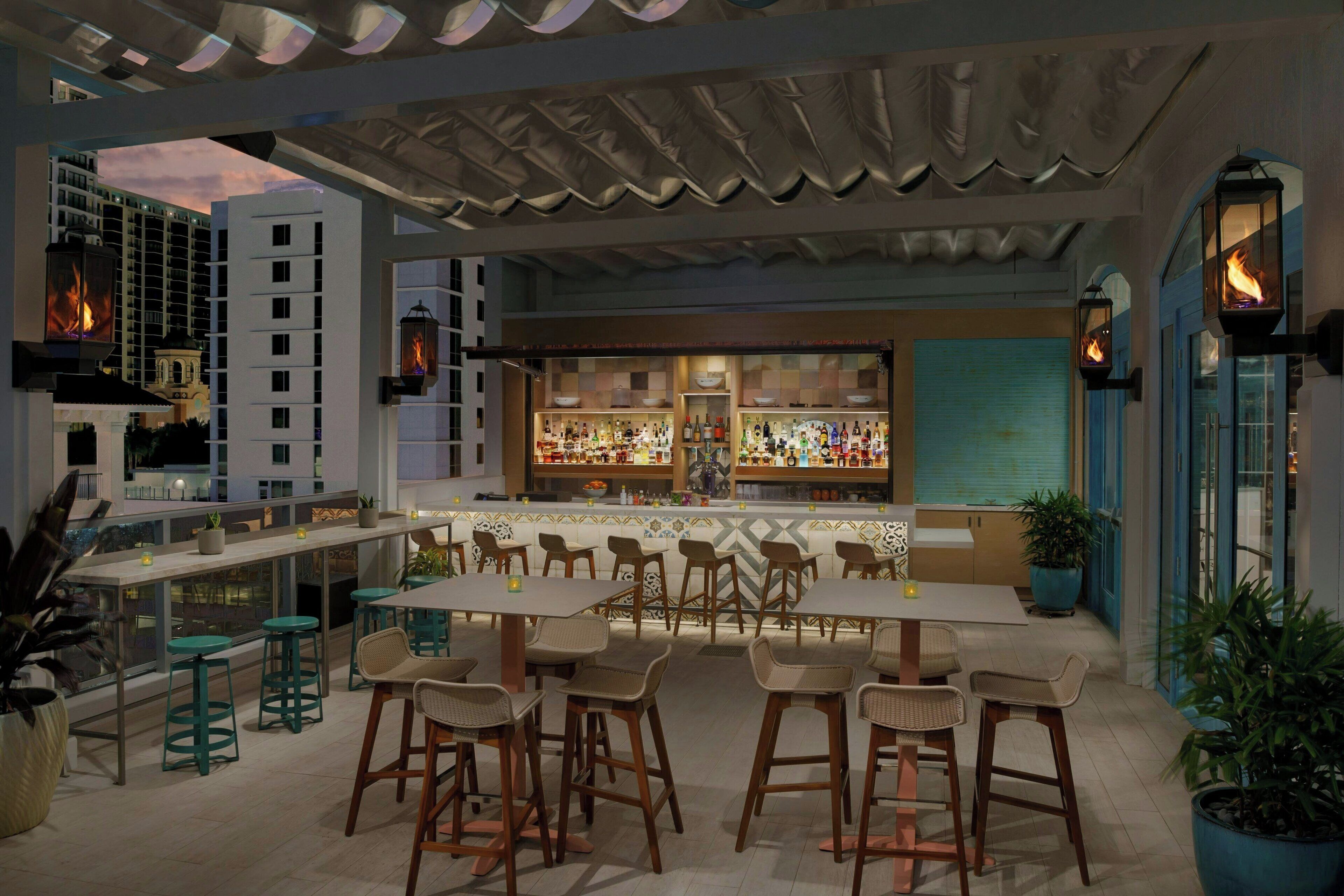 Um bar no terraço com um balcão comprido, vários bancos e vista para a paisagem urbana.