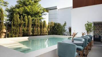 Uma área de piscina moderna ao ar livre, com degraus dando acesso à água, cercada por vegetação exuberante e uma fileira de cadeiras azul-turquesa.