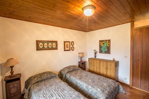 Quarto com duas camas, teto de madeira, luminárias fixadas na parede, um armário de madeira e uma porta.