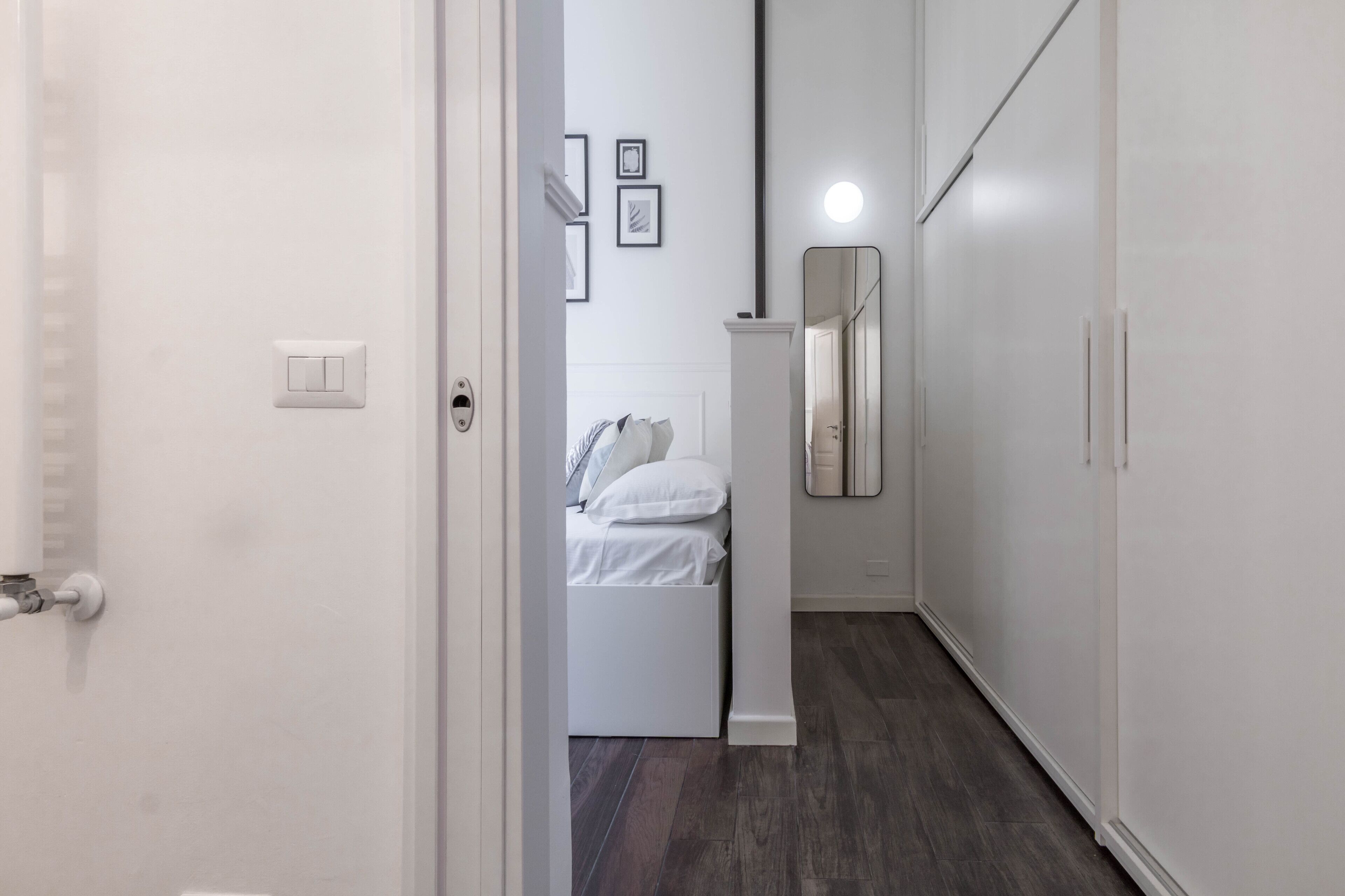 Um corredor estreito com uma cama ao fundo, um guarda-roupa branco e um espelho na parede.