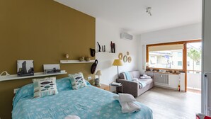 Quarto com cama, sofá, mesinha e janela com vista para prédios.
