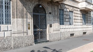 Fachada de edifício com uma grande entrada em arco, paredes de pedra e grades de metal decorativas nas janelas.