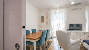 Uma sala de estar moderna com uma mesa de jantar de madeira, cadeiras azuis, um sofá branco e uma televisão.