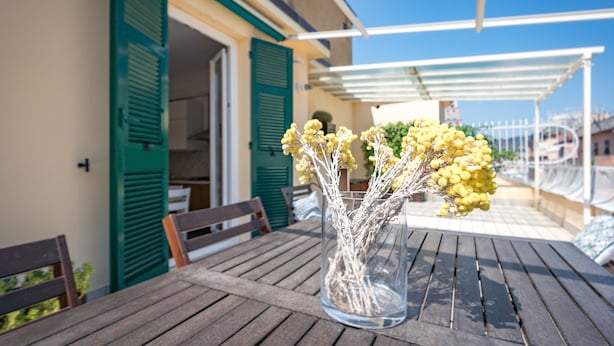 Uma mesa de madeira com um vaso de vidro transparente contendo flores amarelas, colocada em um pátio externo com uma pérgola branca acima.