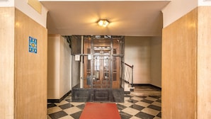 Um saguão de hotel com piso xadrez, tapete vermelho e portão de metal que dá acesso a um elevador.